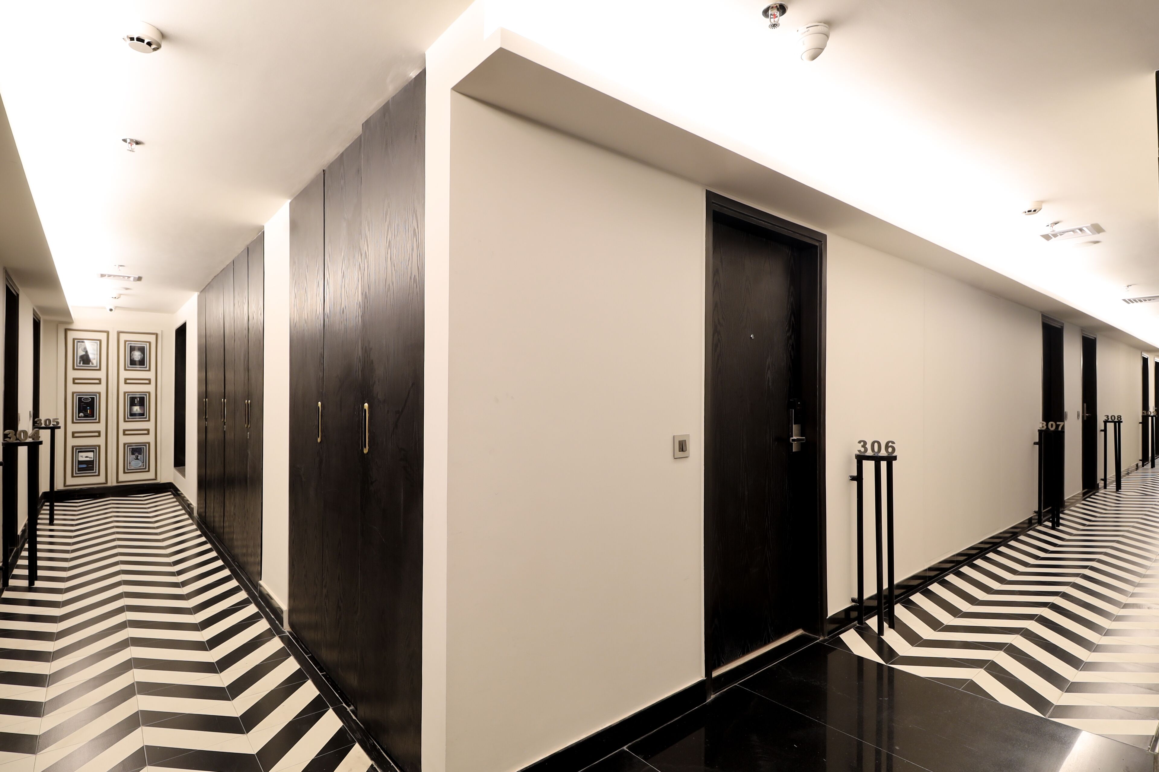 Um corredor moderno com revestimento em padrão Chevron preto e branco, portas de madeira escura e placas com números indicando a localização dos quartos.