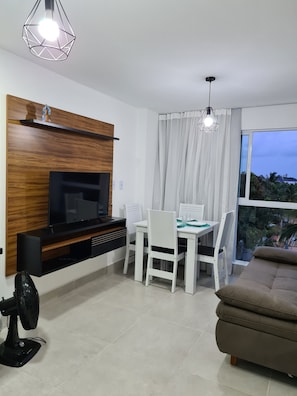 Uma sala de estar moderna com televisão de tela plana, mesa de jantar com cadeiras, sofá e ventilador de teto.