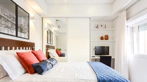Quarto de hotel com cama, escrivaninha com computador, televisão e estante de livros.