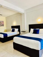Duas camas de solteiro com cabeceiras pretas e roupa de cama branca, uma mesinha de cabeceira e uma luminária fixada na parede.