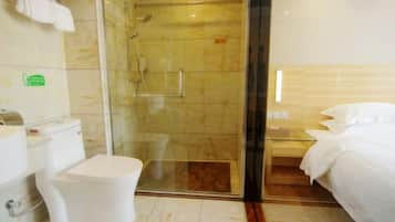 Um banheiro de hotel com chuveiro em cabine de vidro, vaso sanitário e cama.