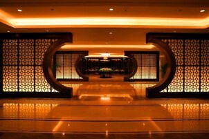 Um hall de hotel com uma mesa de recepção, uma parede decorativa com padrão geométrico e um chão espelhado.