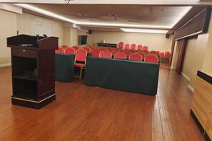 Sala de conferências com fileiras de cadeiras vermelhas, um púlpito e mesas cobertas por toalhas verdes.