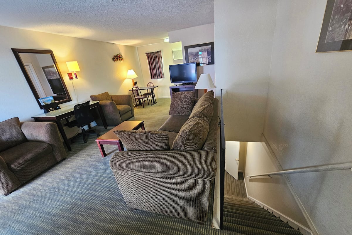 Quarto de hotel com sofá, poltrona, escrivaninha, televisão e escada.
