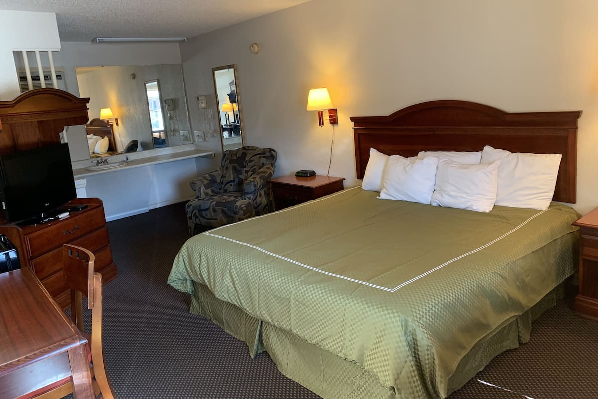 Quarto de hotel com cama, poltrona, escrivaninha com televisão, luminária e espelho.