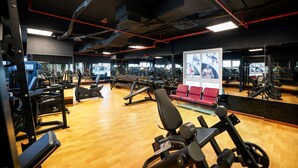 Uma academia com diversos equipamentos de exercício, piso de madeira e um grande painel de espelhos.