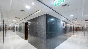 Corredor moderno de hotel com parede de mármore polido, um elevador de vidro e sinalização de saída visível.