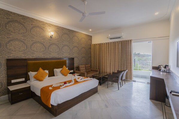 Quarto de hotel com uma cama grande, área de estar com mesa e cadeiras, varanda com cortinas e ventilador de teto.