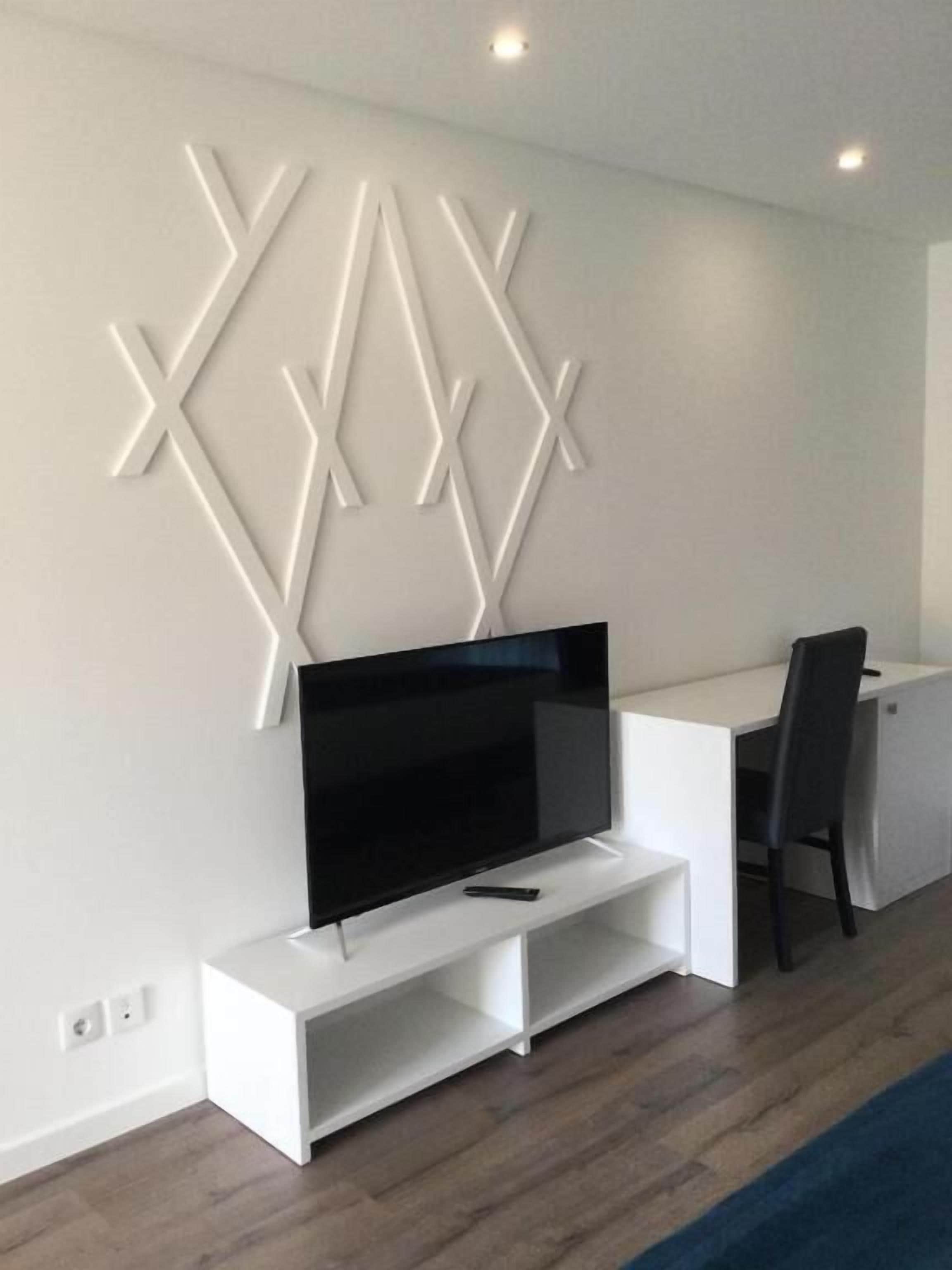 Uma sala de estar moderna com uma TV de tela plana sobre um móvel branco, uma escrivaninha com uma cadeira e um painel decorativo na parede.