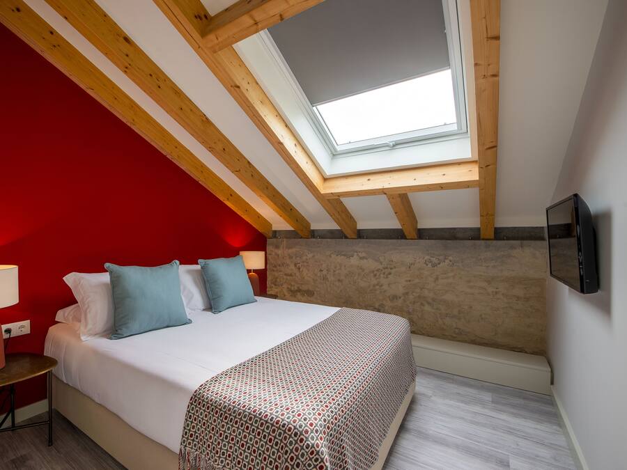 Quarto com teto inclinado, claraboia, cama com travesseiros, mesa de cabeceira com abajur e manta estampada.