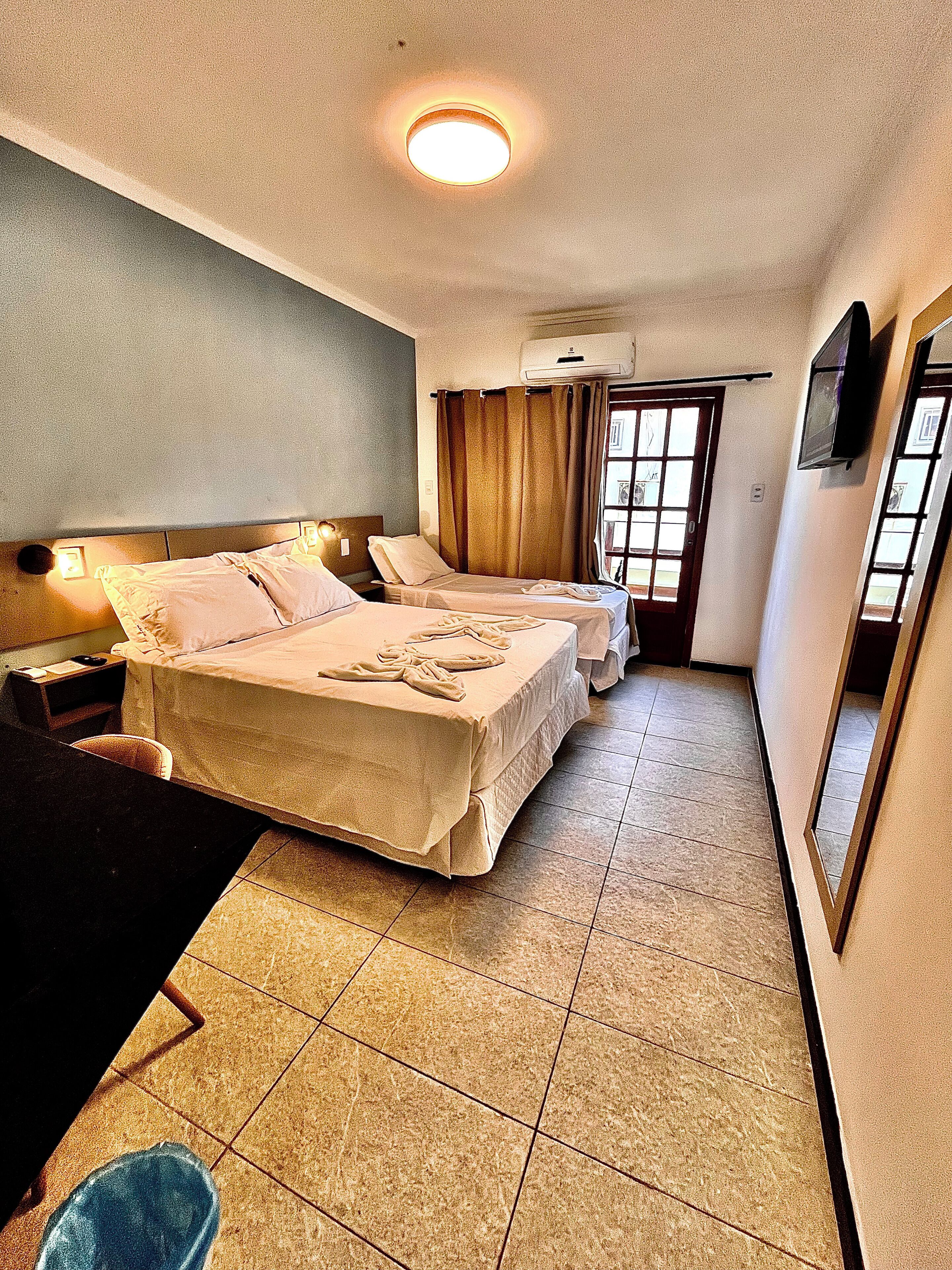 Quarto de hotel com duas camas, uma mesa, uma cadeira, televisão e janela com cortinas.