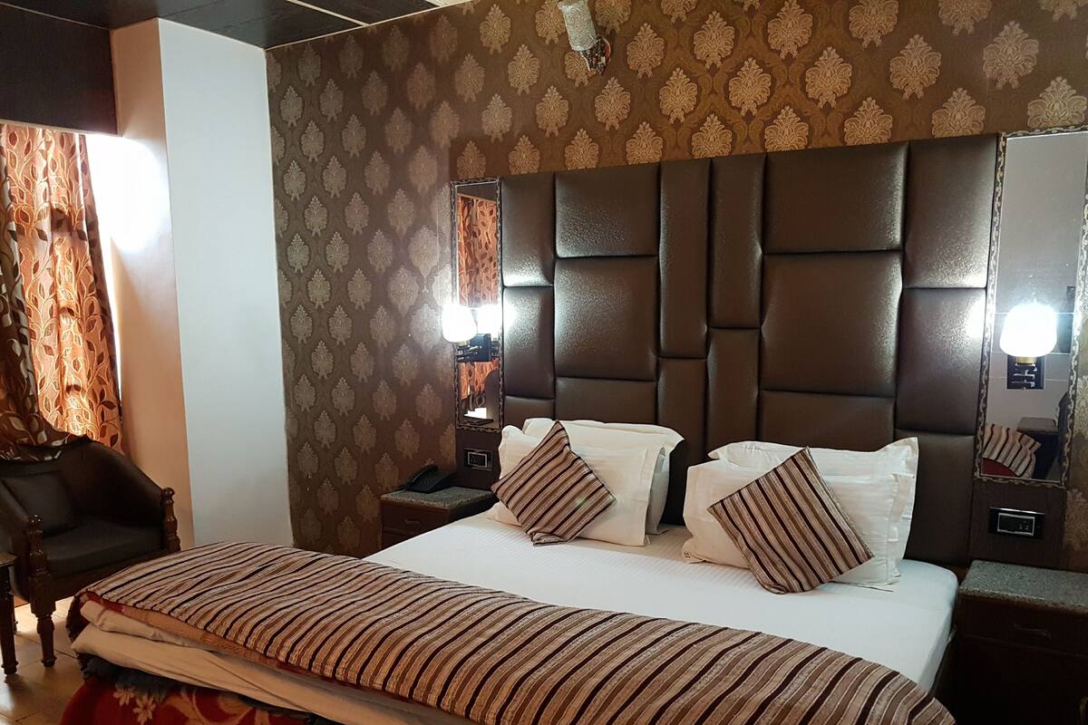 Quarto de hotel com uma cama grande, cabeceira, mesa de cabeceira, luminária, espelho e uma cadeira.