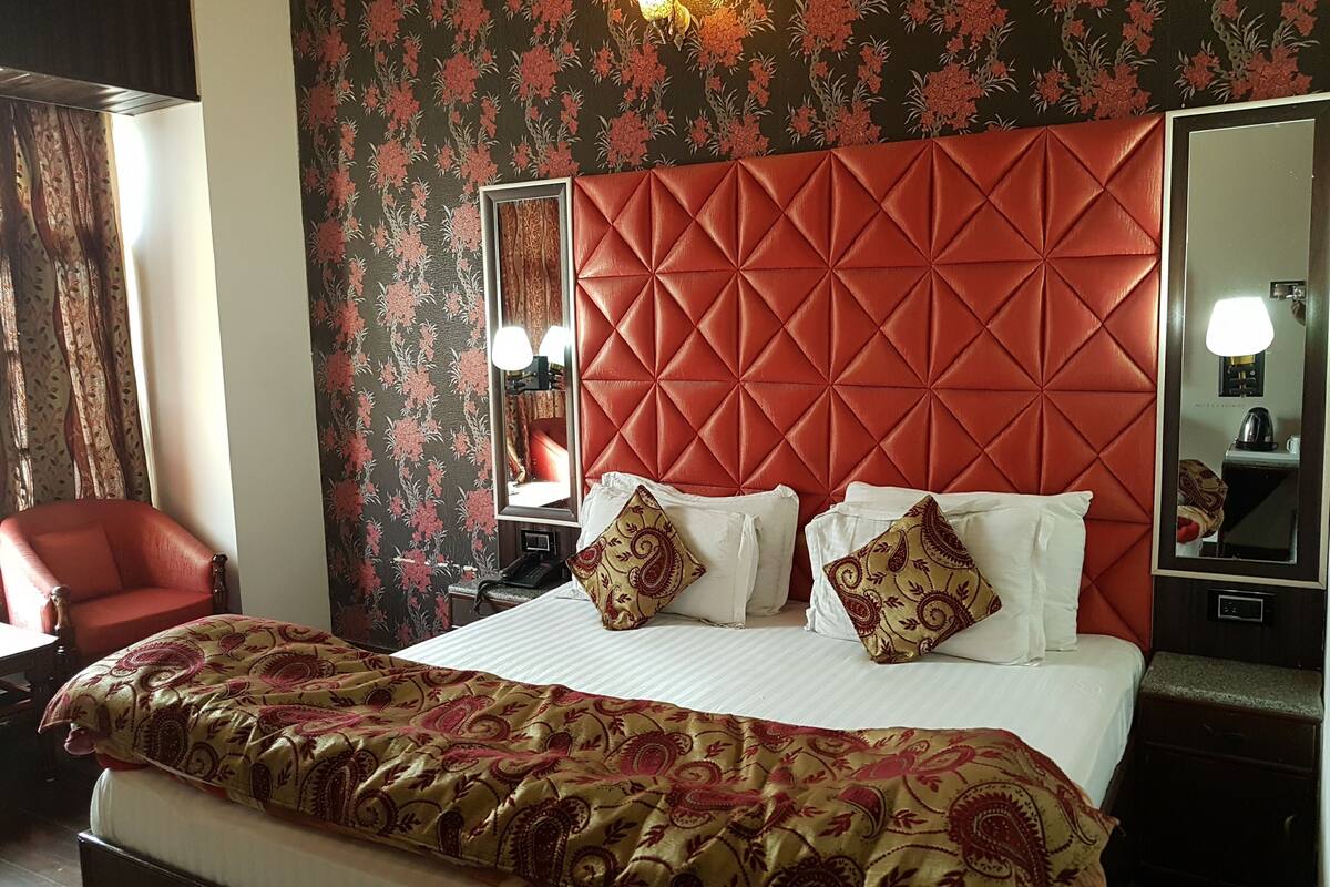 Quarto de hotel com uma cama grande, cabeceira vermelha, papel de parede floral, um espelho e uma poltrona vermelha.