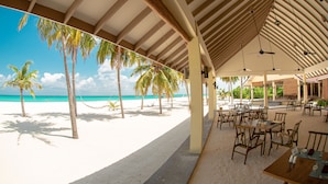 Restaurante à beira-mar com mesas ao ar livre, com vista para uma praia de areia e águas azul-turquesa.