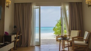 Quarto com escrivaninha, cadeiras, uma pequena mesa com uma cesta de frutas e vista para a praia através de uma porta de vidro.
