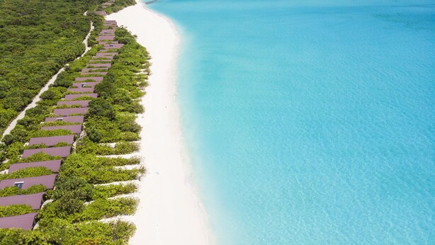 Vista aérea de um resort na praia, com fileiras de quartos ao longo da costa, águas turquesa cristalinas e uma praia de areia branca.