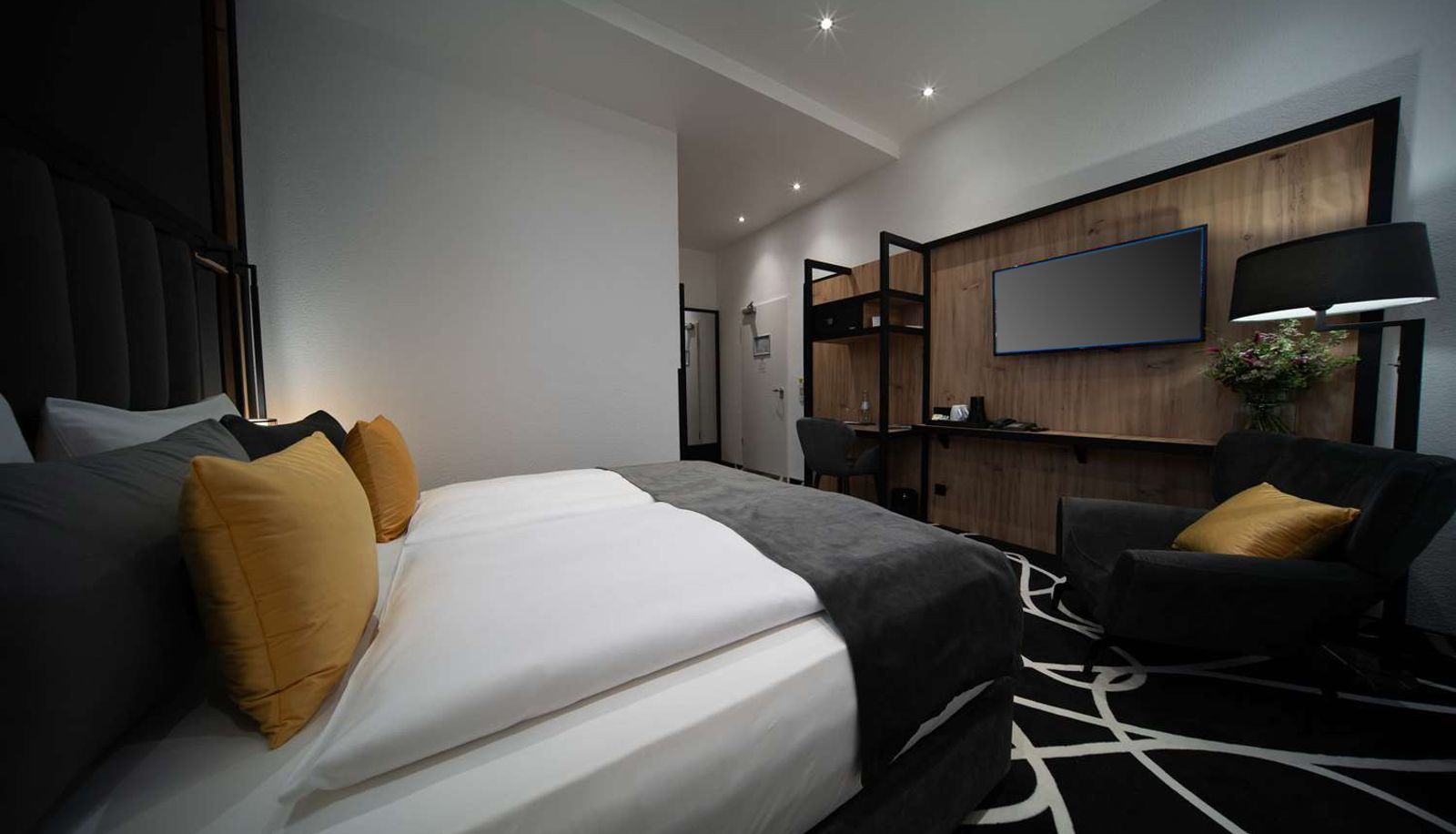 Quarto de hotel moderno com uma cama grande, uma mesa de escritório, uma cadeira e uma televisão de tela plana na parede.