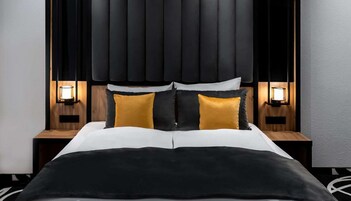 Uma cama com edredom preto e branco, duas mesas de cabeceira pretas com abajures e uma cabeceira preta com ripas verticais.