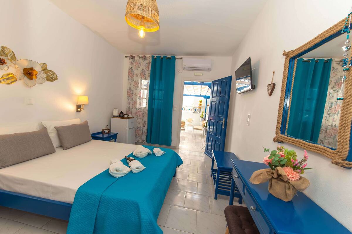 Quarto de hotel com cama, secretária, espelho, televisão presa à parede e banheiro visível por uma porta aberta.