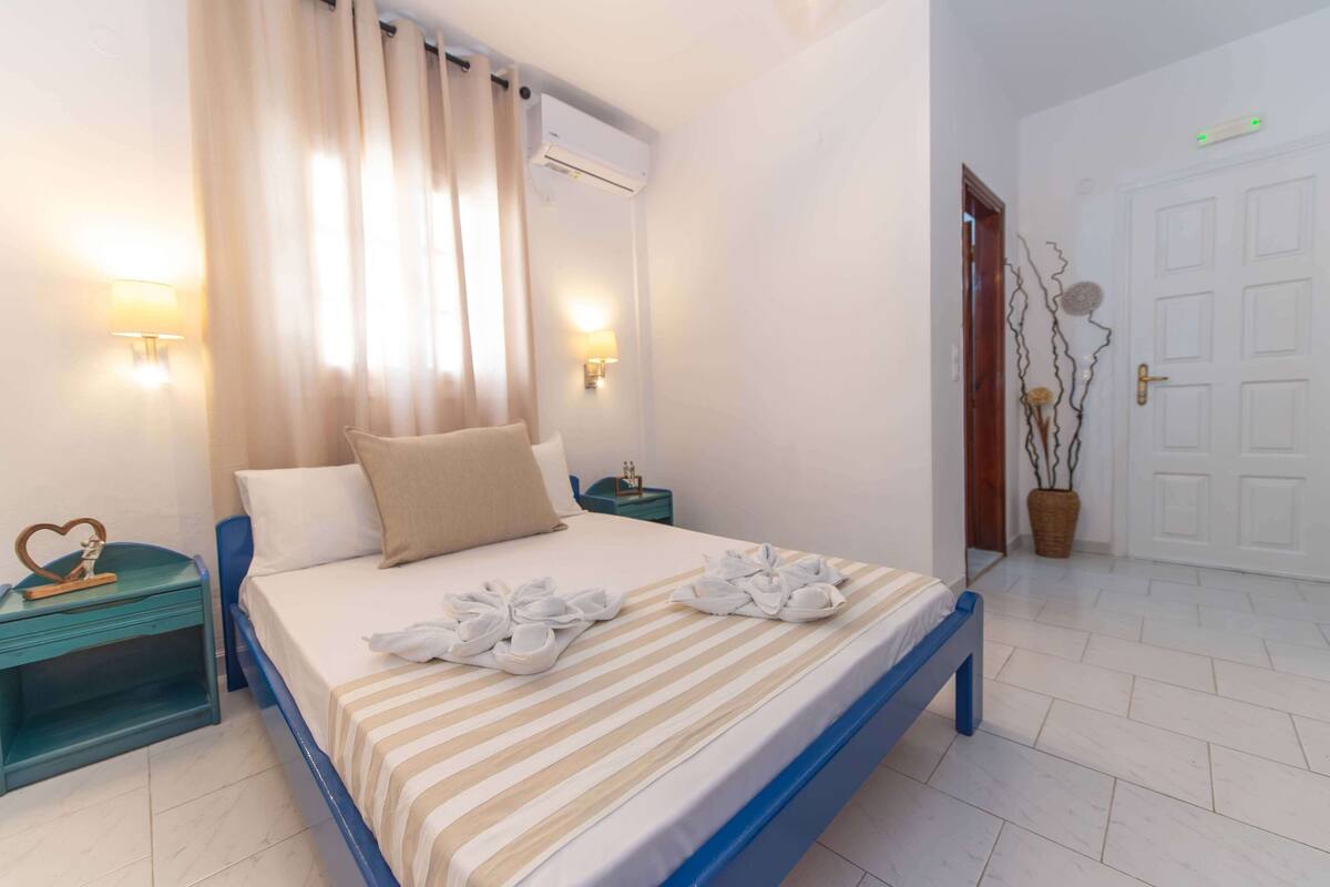 Quarto de hotel com cama, duas mesas de cabeceira, um criado-mudo, ar-condicionado na parede, uma porta e um vaso de plantas.
