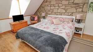 Quarto com parede de pedra, cama com roupa de cama florida, mesa de cabeceira de madeira, televisão e janela com persianas.