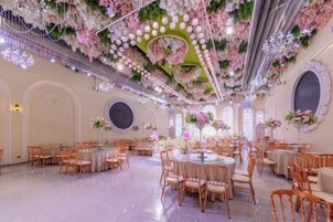 Uma sala de banquetes com mesas redondas e cadeiras, decorada com arranjos florais e luminárias pendentes.