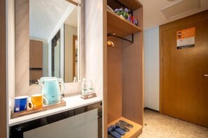 Quarto de hotel com cafeteira, garrafas de água, espelho, prateleira com chinelos e porta de madeira.