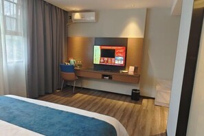 Quarto de hotel com cama, escrivaninha com computador, televisão e janela com cortinas.