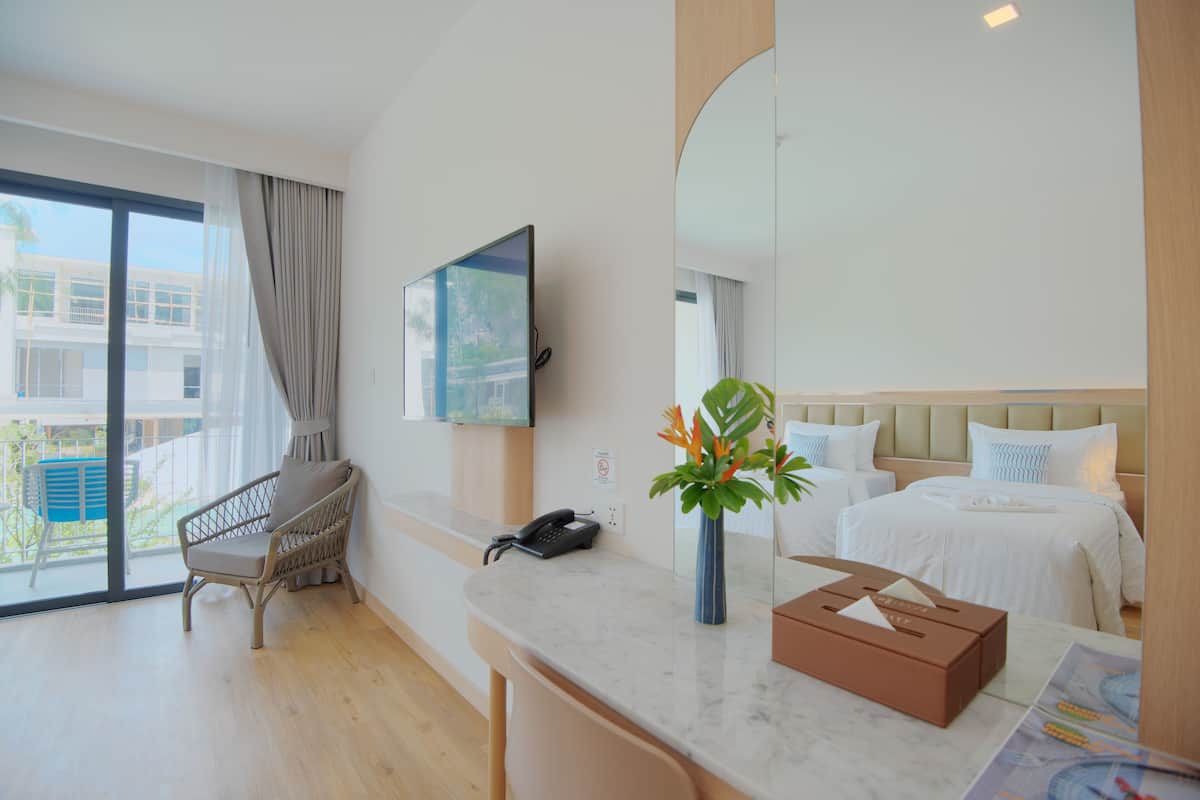 Quarto de hotel com uma janela grande, cama, escrivaninha com telefone, cadeira e vaso com flores.