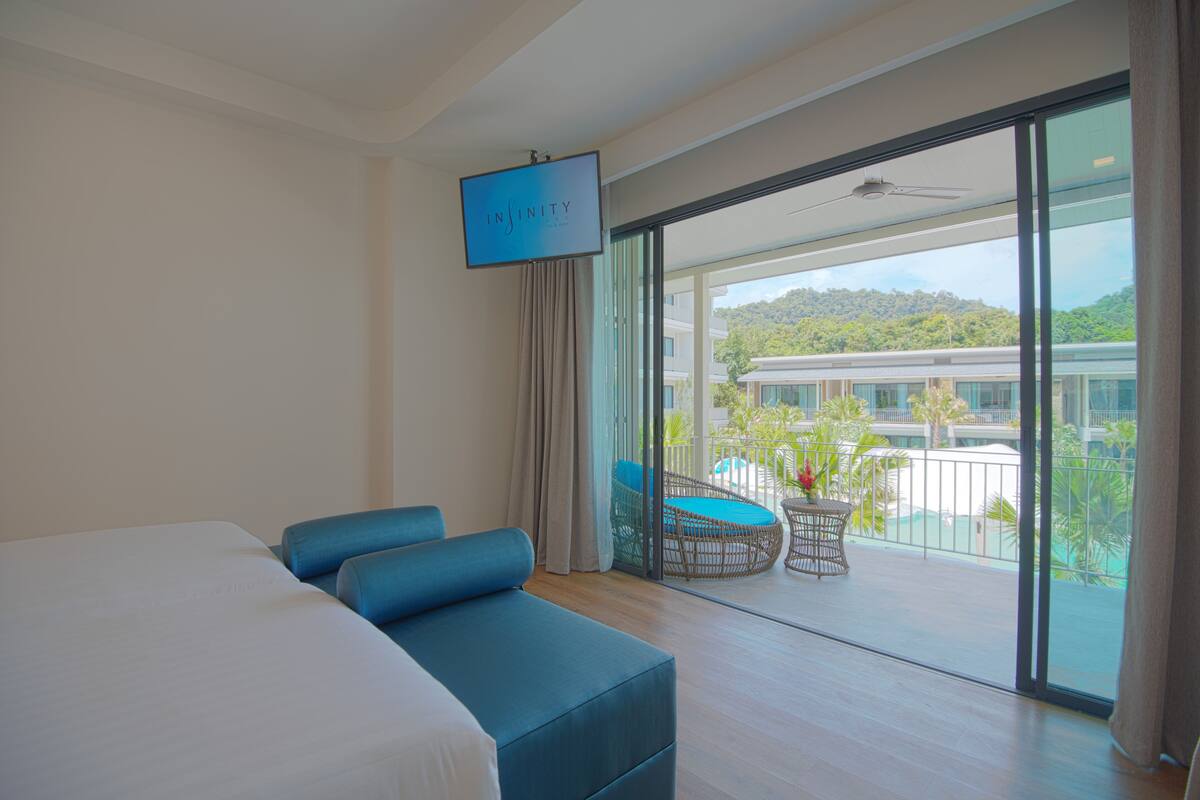 Quarto de hotel moderno com uma cama grande, um sofá azul, uma televisão e uma varanda com vista para um edifício e vegetação.