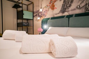 Uma cama bem arrumada com toalhas brancas dobradas, um painel de cabeceira verde e uma parede com um quadro floral.