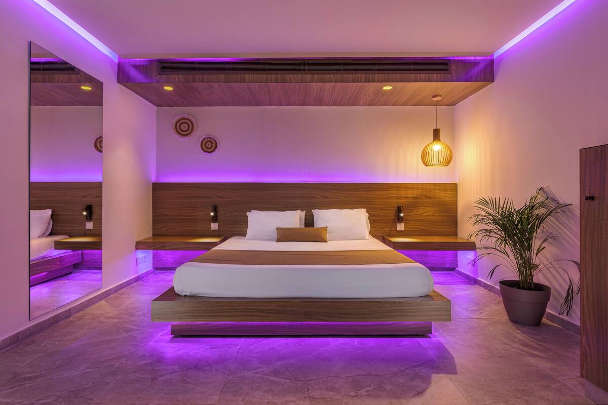 Quarto moderno com cama grande, cabeceira de madeira, iluminação ambiente e um vaso com planta.