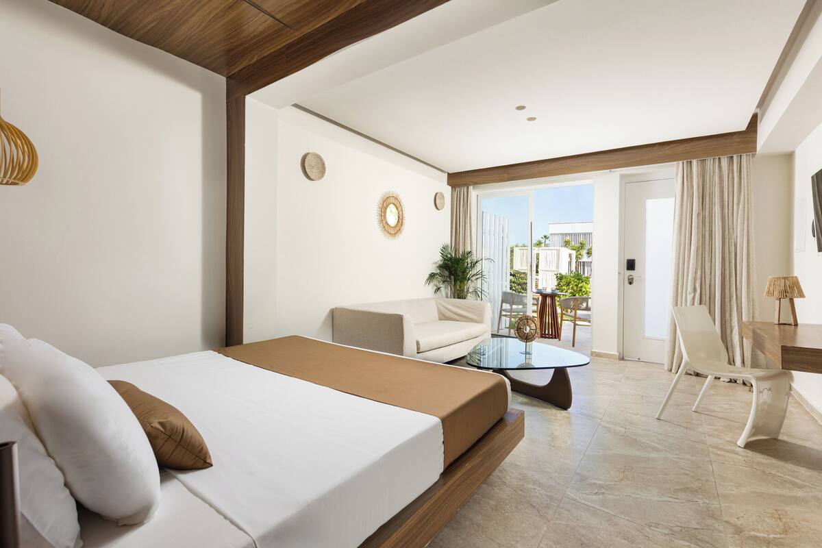 Quarto de hotel moderno com cama, sofá, mesa de centro de vidro e vista para o exterior.