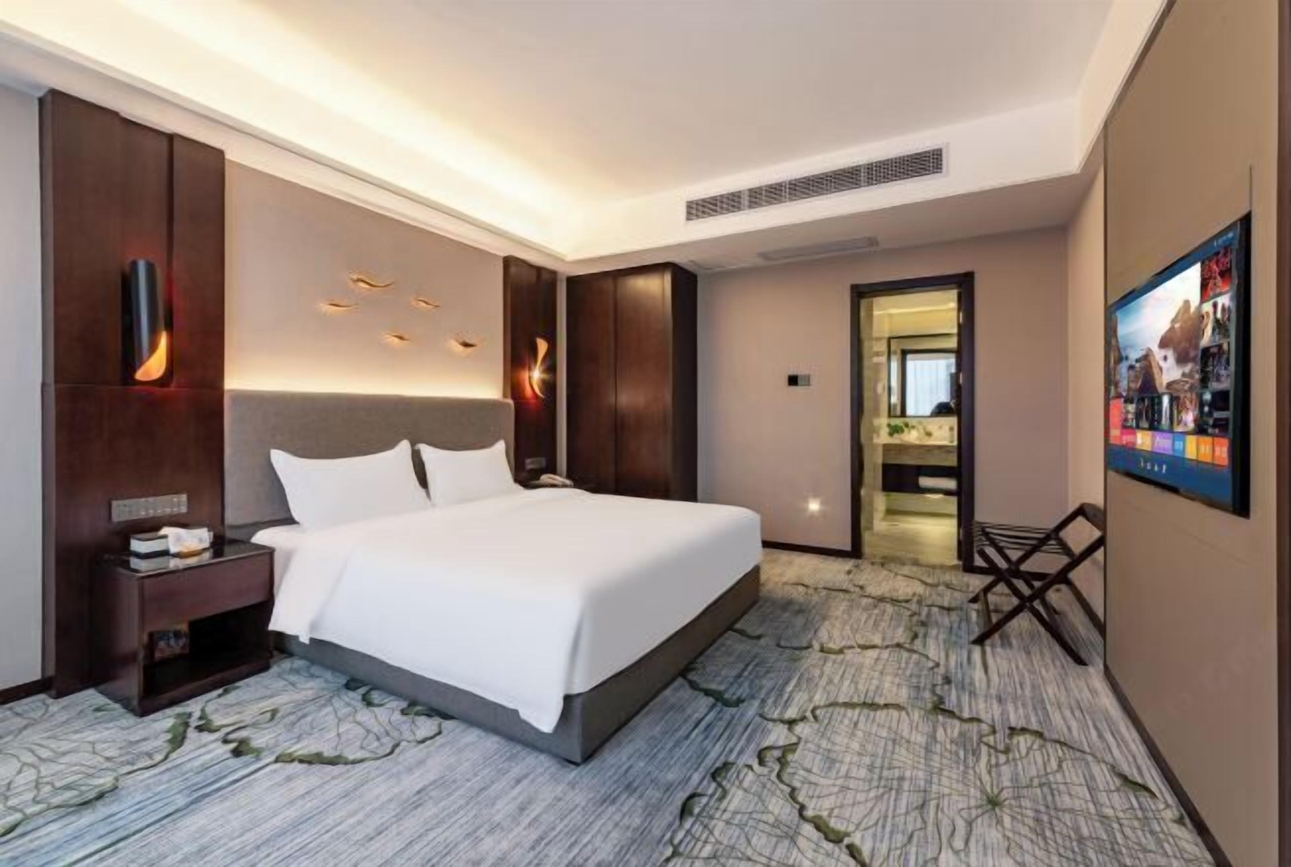 Quarto de hotel com uma cama grande, mesa de cabeceira, televisão e banheiro visível por uma porta aberta.