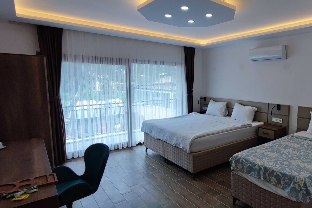 Quarto de hotel com duas camas, uma escrivaninha, uma cadeira e uma janela grande com cortinas.