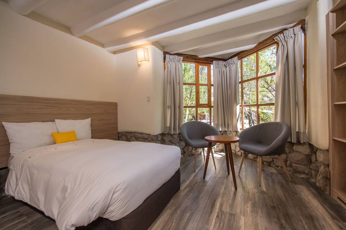 Quarto com uma cama grande, duas cadeiras, uma mesinha, uma parede de pedra e uma janela com cortinas.
