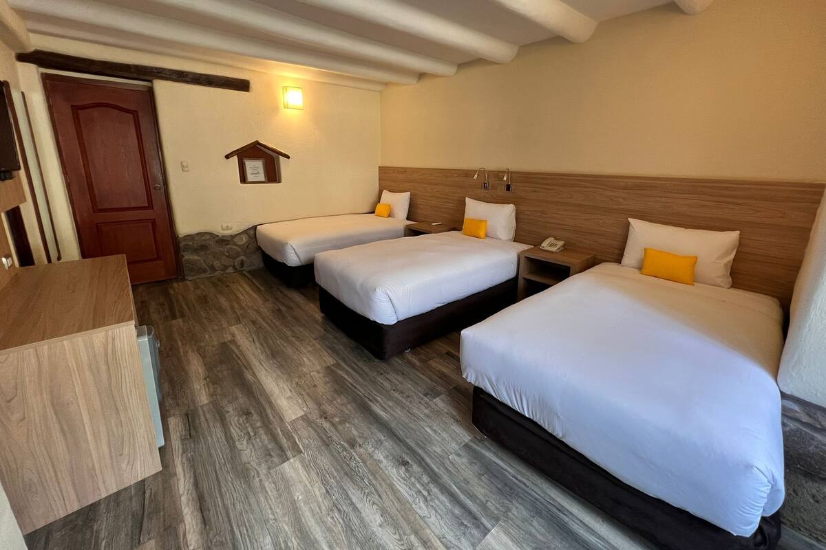 Quarto de hotel com três camas de solteiro, uma escrivaninha de madeira, um pequeno armário de madeira e uma parede de pedra.