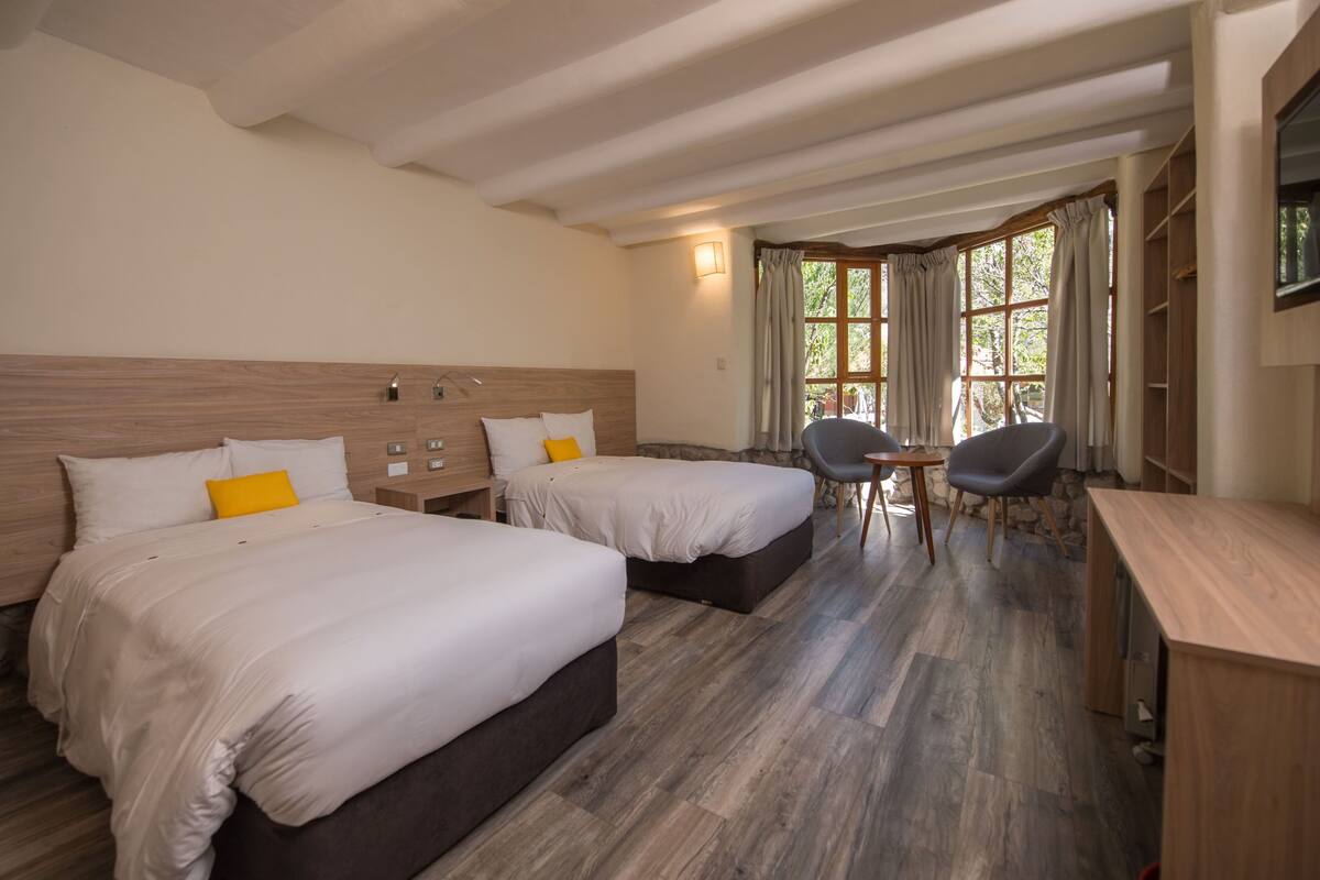Quarto de hotel com duas camas, uma escrivaninha, uma cadeira e uma janela com cortinas.