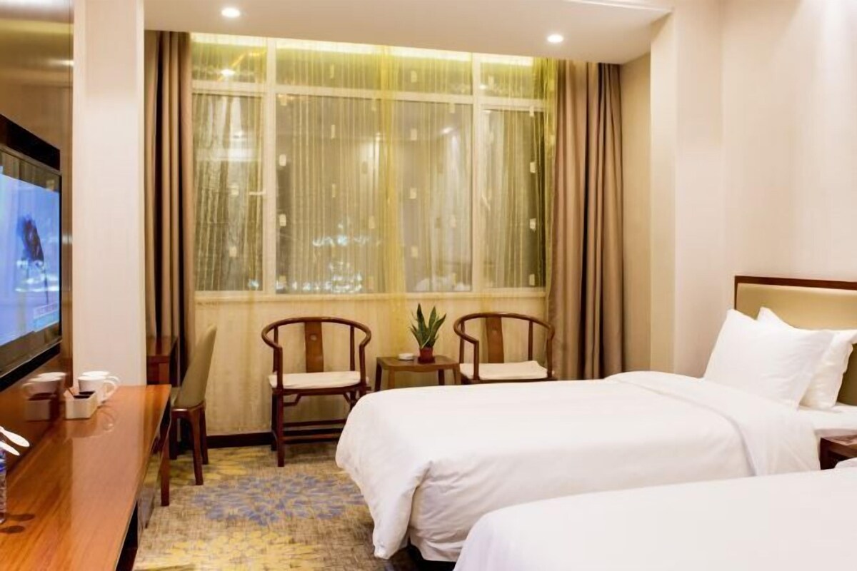 Quarto de hotel com duas camas, uma televisão, uma mesa com cadeira e uma janela com cortinas.