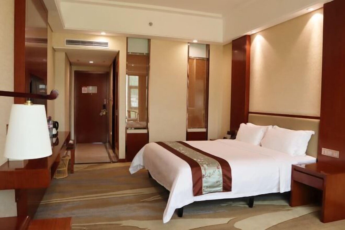 Quarto de hotel com cama grande, mesa de cabeceira, abajur e vista para o corredor.