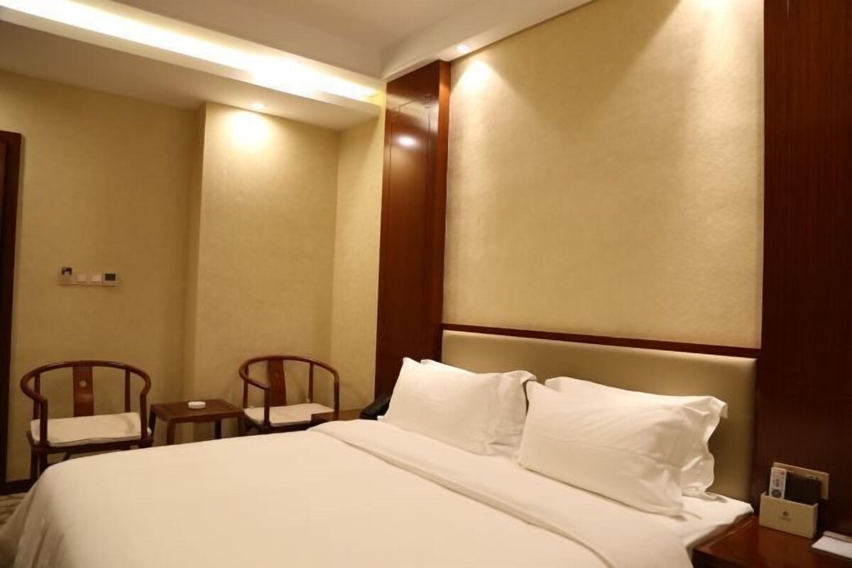 Quarto de hotel com uma cama grande, duas cadeiras de madeira, uma mesinha e uma mesa de cabeceira com abajur.