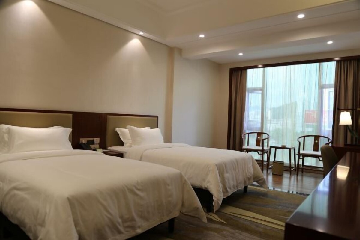 Quarto de hotel com duas camas, uma escrivaninha, cadeiras e uma janela grande com cortinas.
