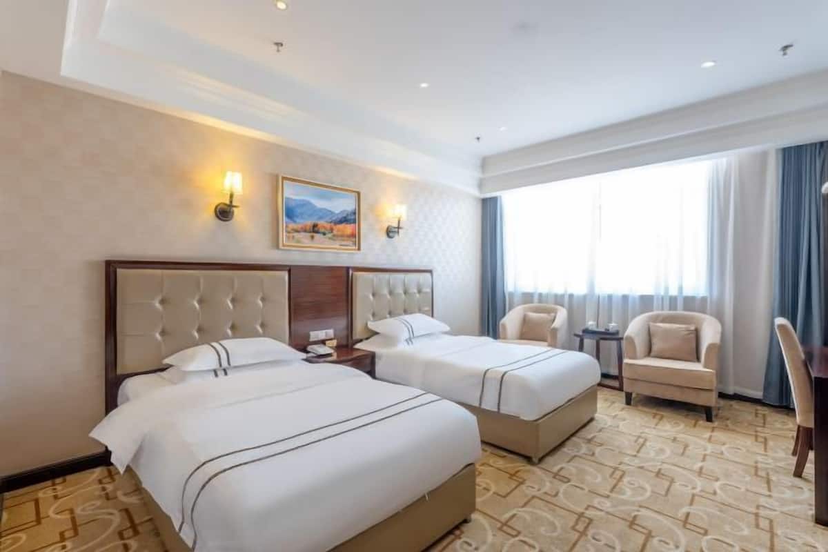Quarto de hotel com duas camas, uma escrivaninha, uma cadeira e uma janela grande.