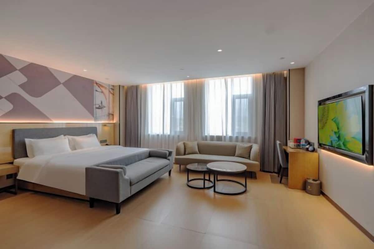 Quarto de hotel moderno com cama, sofá, duas mesinhas, escrivaninha e televisão.