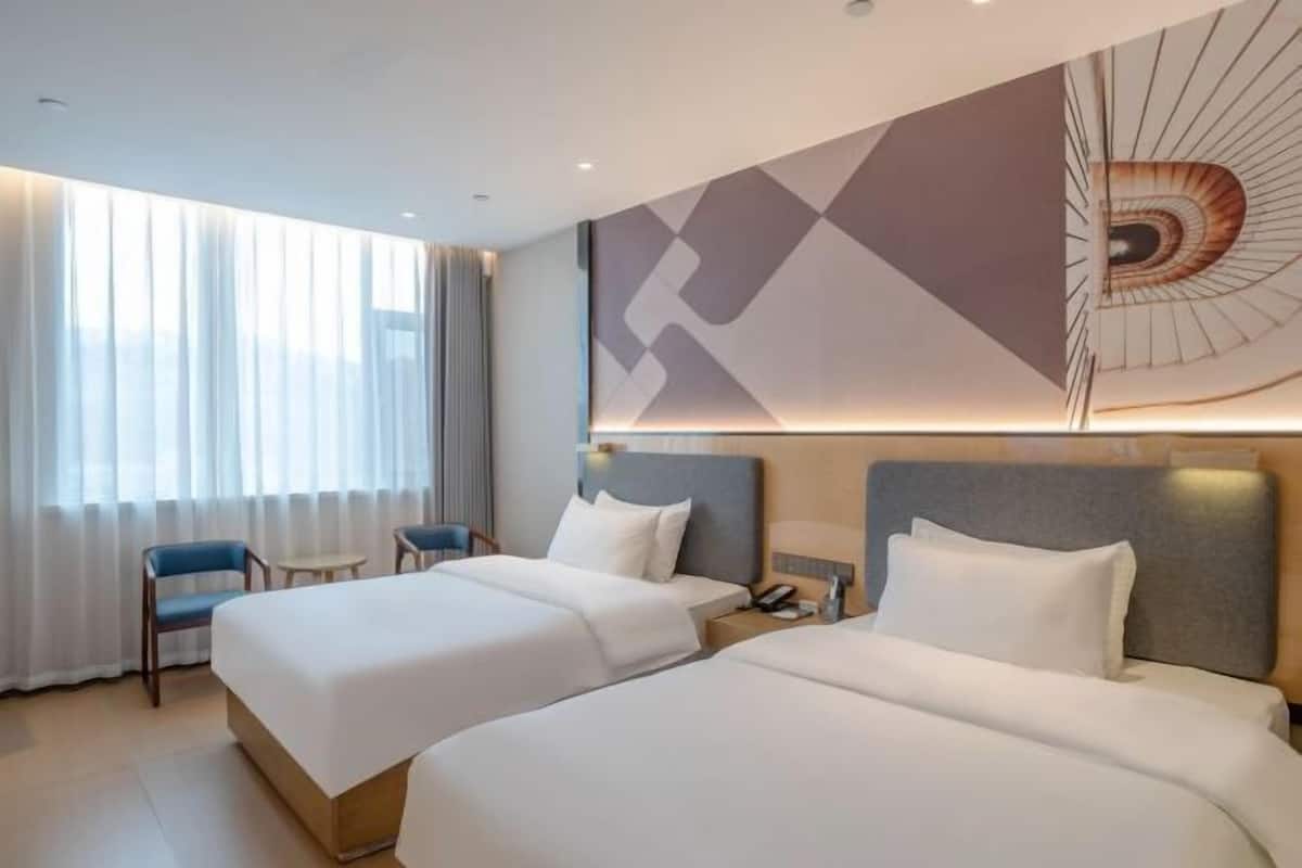 Quarto de hotel com duas camas, uma escrivaninha, uma cadeira e uma janela grande com cortinas.