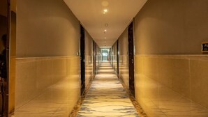 Um corredor comprido e estreito com carpete estampado, paredes em tom bege claro e iluminação embutida no teto.