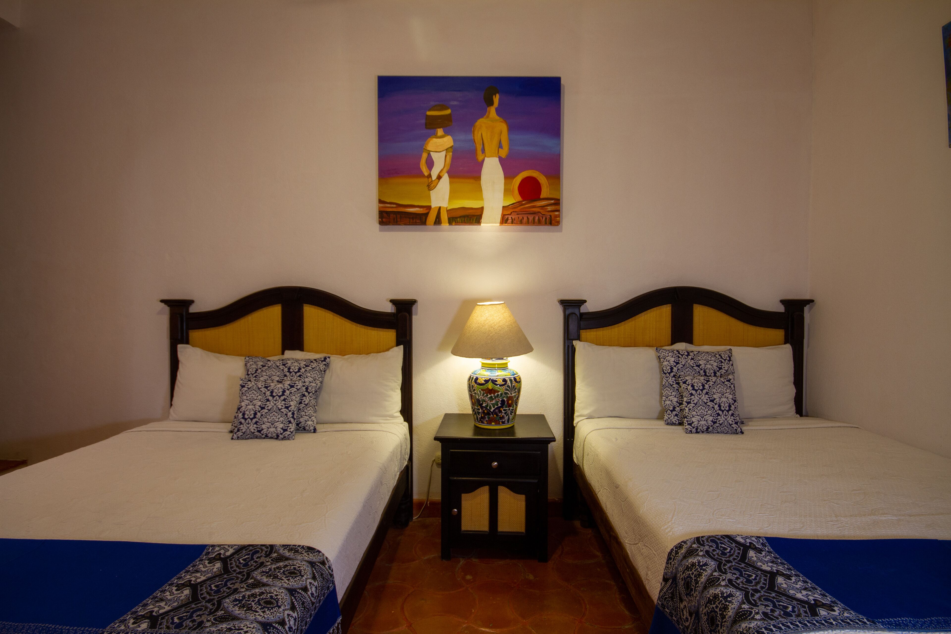 Quarto de hotel com duas camas, uma mesa de cabeceira com abajur, almofadas estampadas e um quadro na parede.