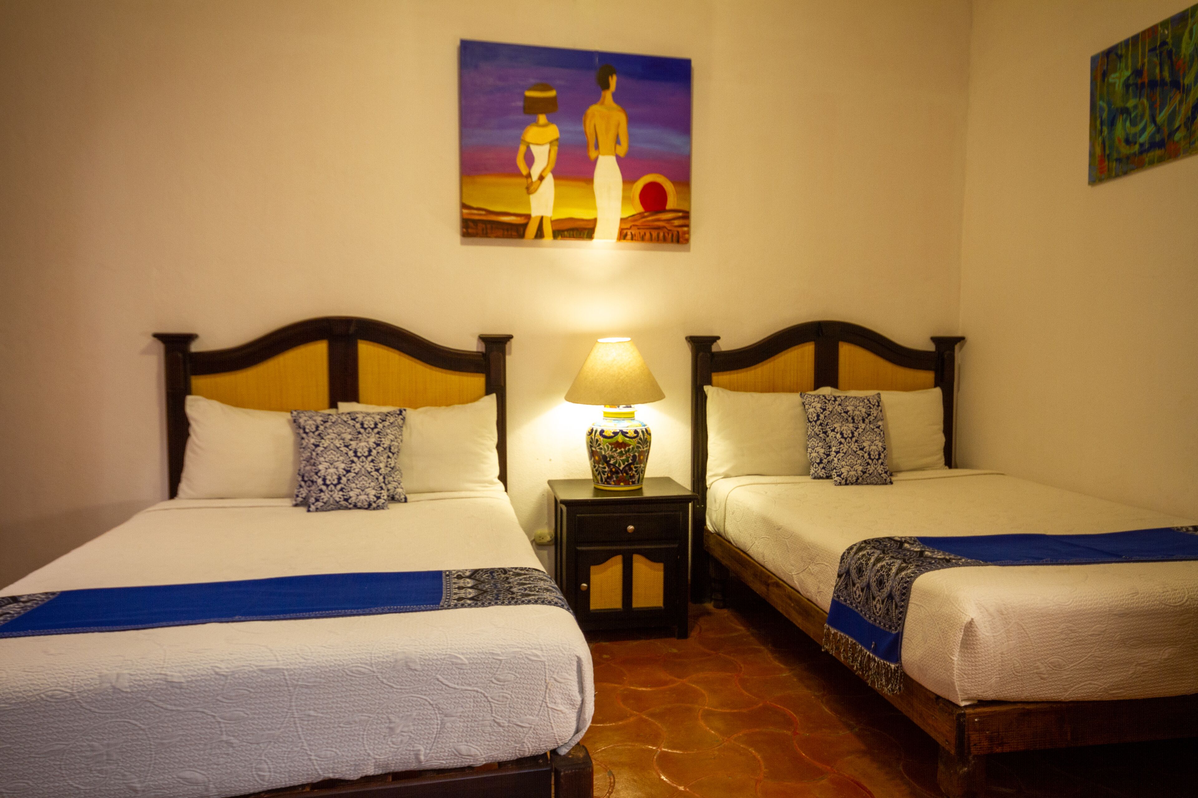 Quarto de hotel com duas camas, cada uma com cabeceira de madeira, roupa de cama branca e manta azul. Há uma mesa de cabeceira com abajur entre as camas, e um quadro com a imagem de duas figuras na parede acima das camas.