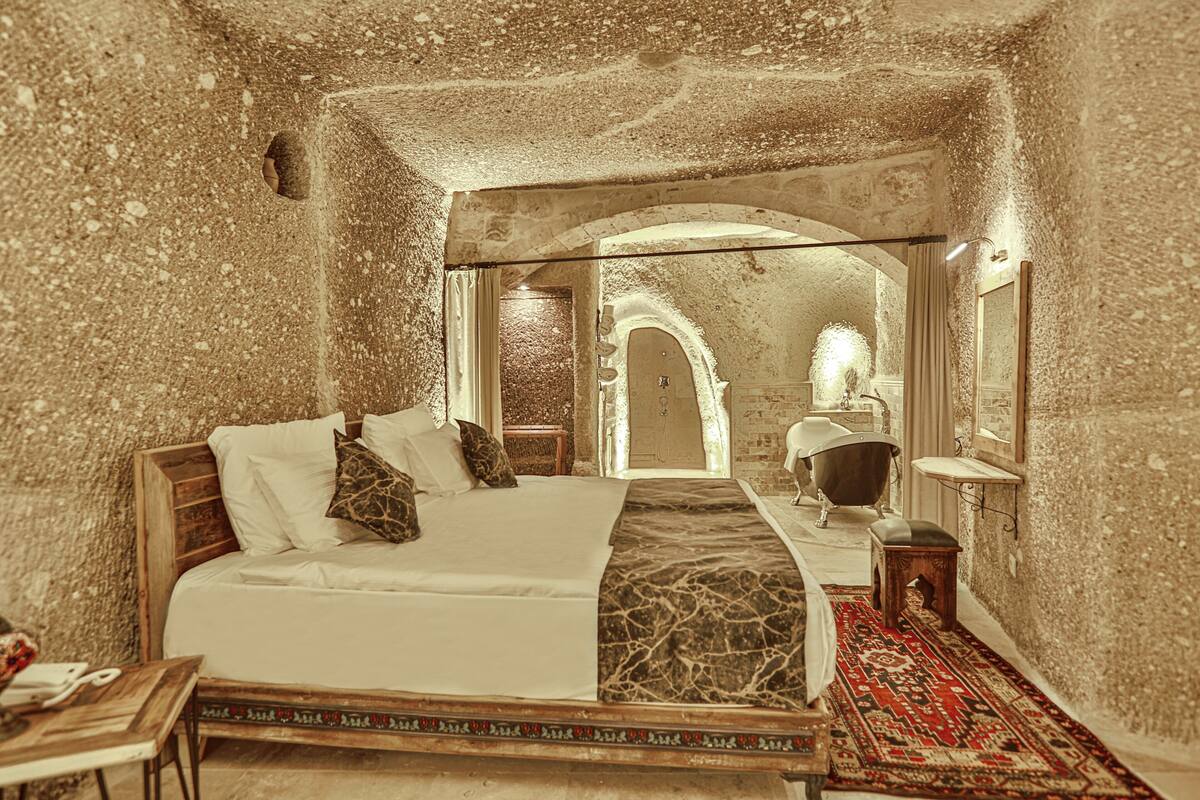 Quarto com cama grande, mesa de cabeceira de madeira, tapete vermelho estampado, banheira antiga e parede de pedra.