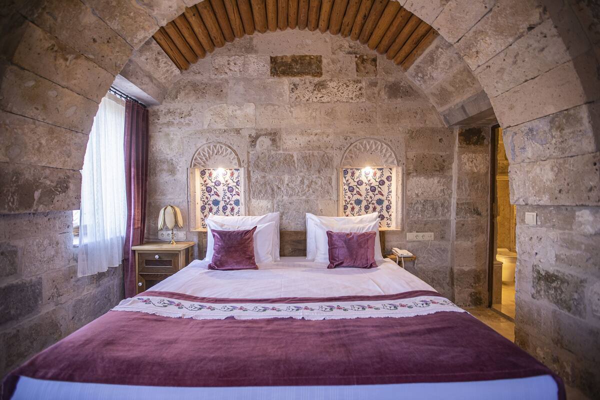 Quarto com paredes de pedra, uma cama grande, duas mesas de cabeceira e teto de madeira.